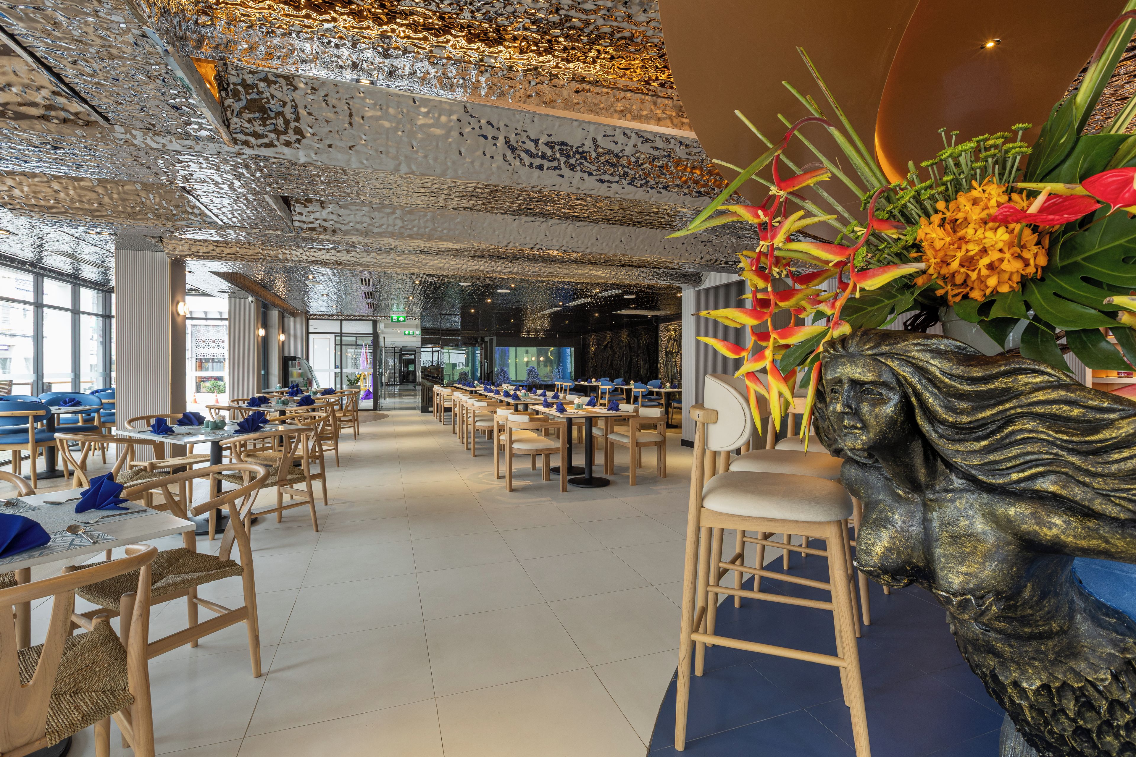 Restaurante com várias mesas e cadeiras, parede decorativa com uma escultura, e vista para o exterior através de grandes janelas.