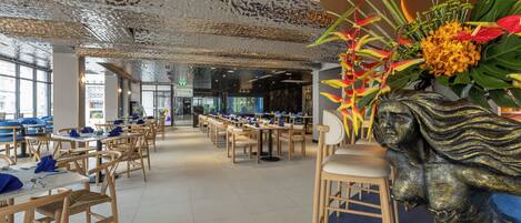 Restaurante com várias mesas e cadeiras, parede decorativa com uma escultura, e vista para o exterior através de grandes janelas.