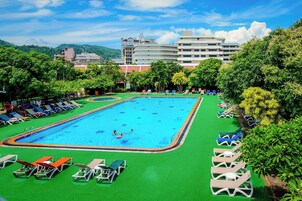 Uma piscina com vários espreguiçadeiras e área do deck verde, cercada por vegetação exuberante e edifícios ao fundo.