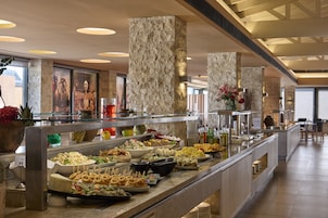 Buffet variado com saladas, sanduíches e aperitivos, além de um pilar de pedra e uma vitrine de vidro.