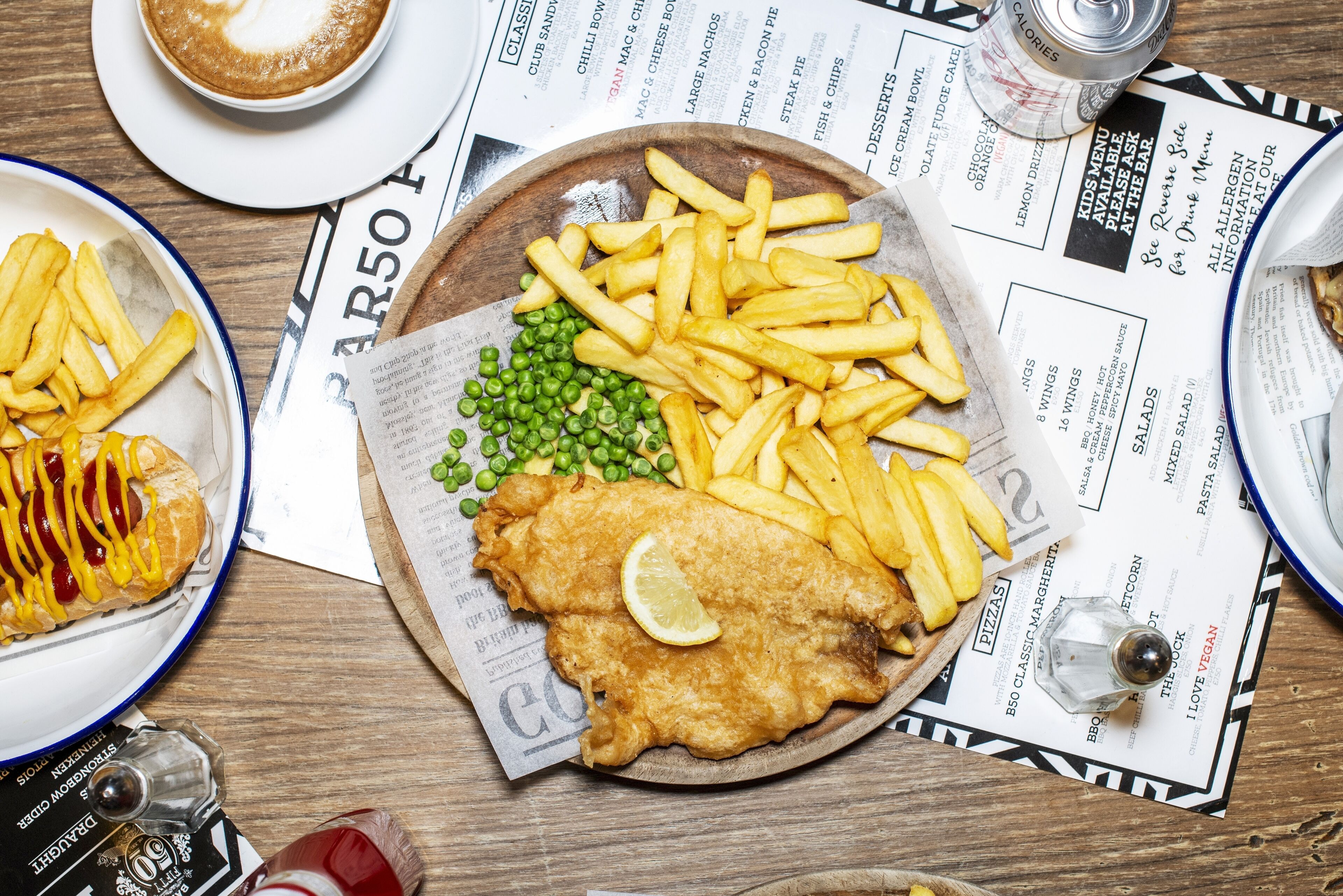 Uma refeição com peixe frito, batatas fritas, ervilhas e um cachorro-quente sobre uma mesa de madeira, com cardápios e um copo de água.