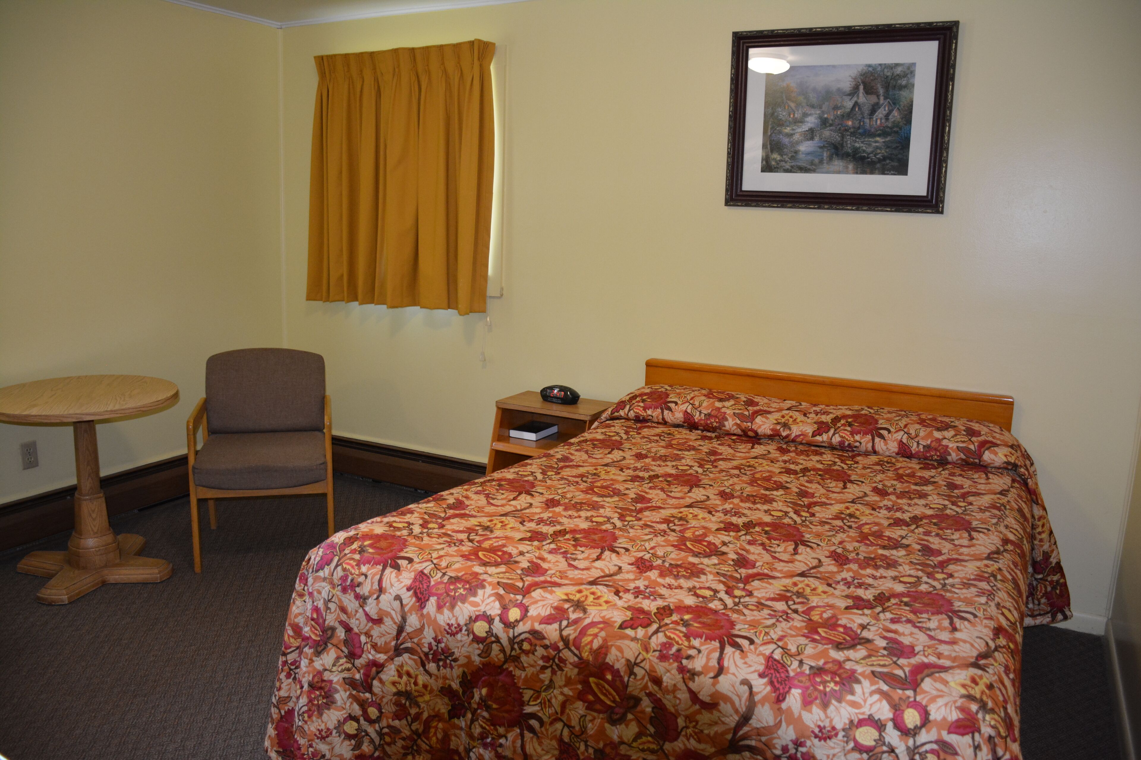 Quarto de hotel com cama, uma cadeira, uma mesinha e um quadro na parede.