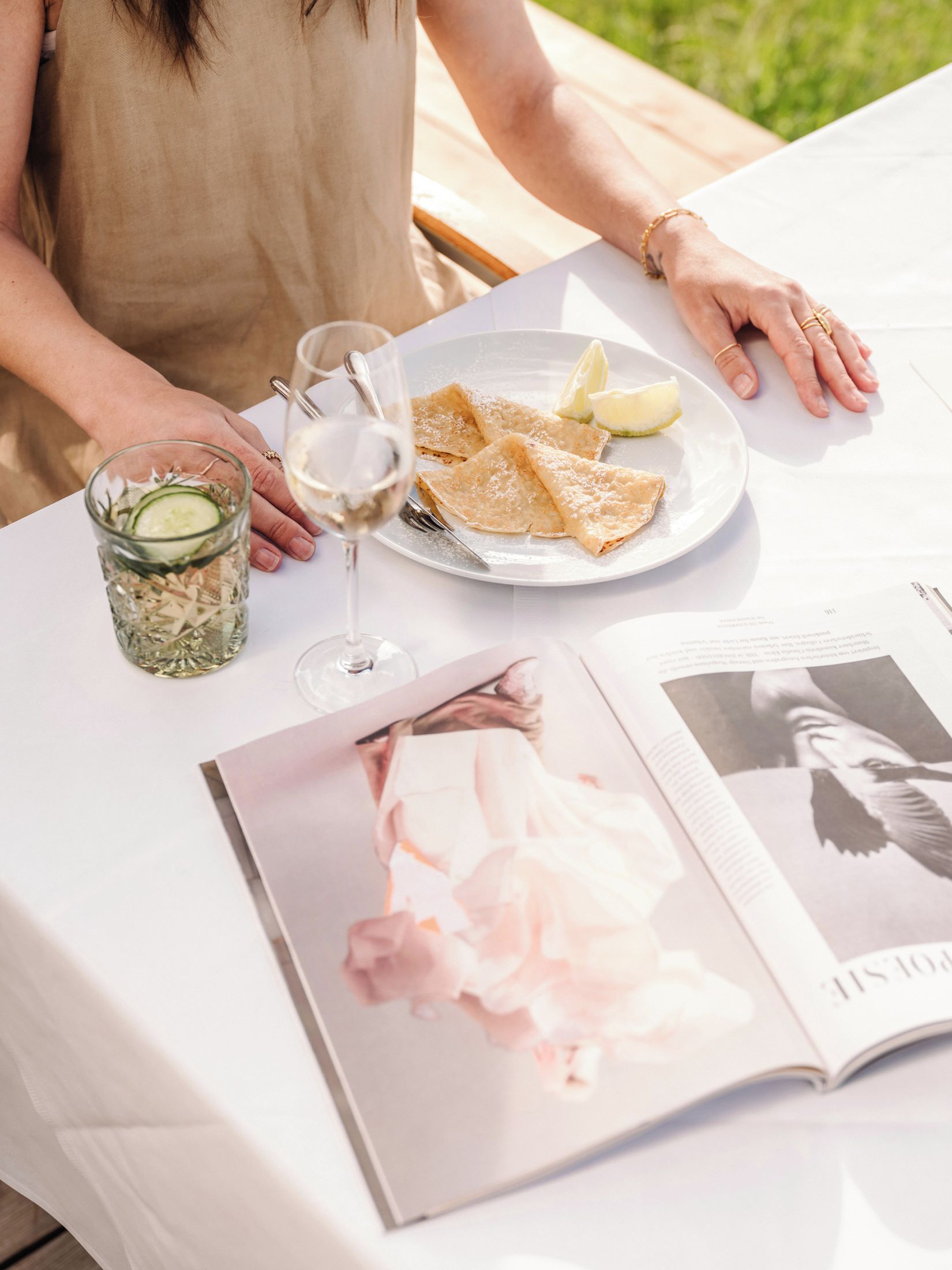 Uma mesa com um prato de comida, um copo de vinho branco e uma revista.