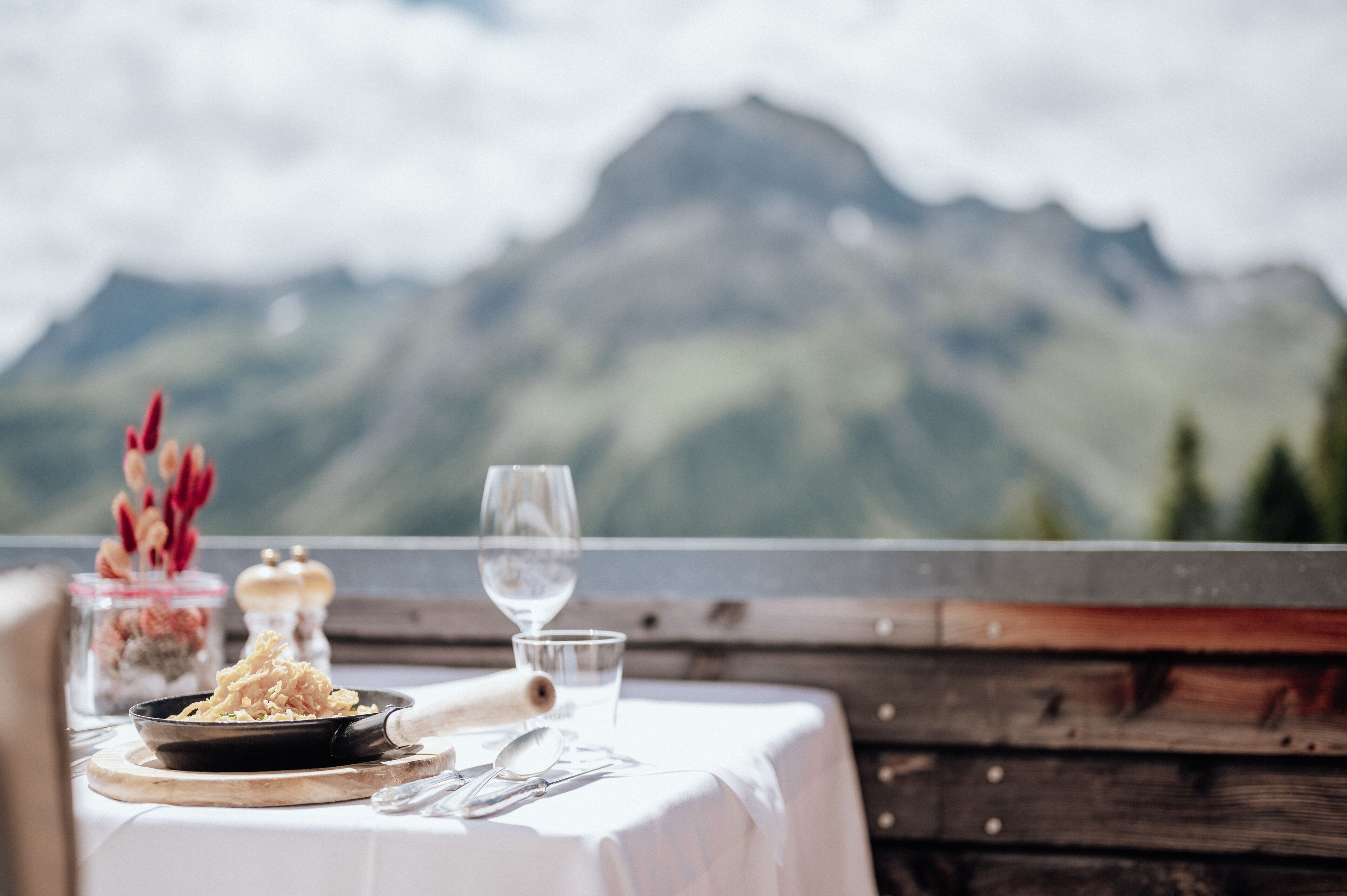 Mesa posta com um prato de comida, um copo de água e uma taça de vinho, com vista para as montanhas ao fundo.