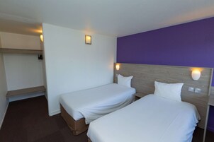 Quarto de hotel com duas camas de solteiro, uma cabeceira de madeira, uma parede com detalhes em roxo e uma pequena prateleira com luz.