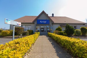 Um hotel chamado Kyriad, com uma área de recepção, telhado telhado e um caminho ladeado por sebes amarelas e verdes.