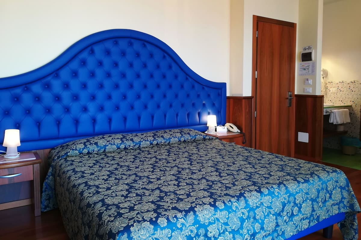 Quarto com cabeceira estofada azul, colcha estampada com flores, abajur e mesa de cabeceira com telefone.
