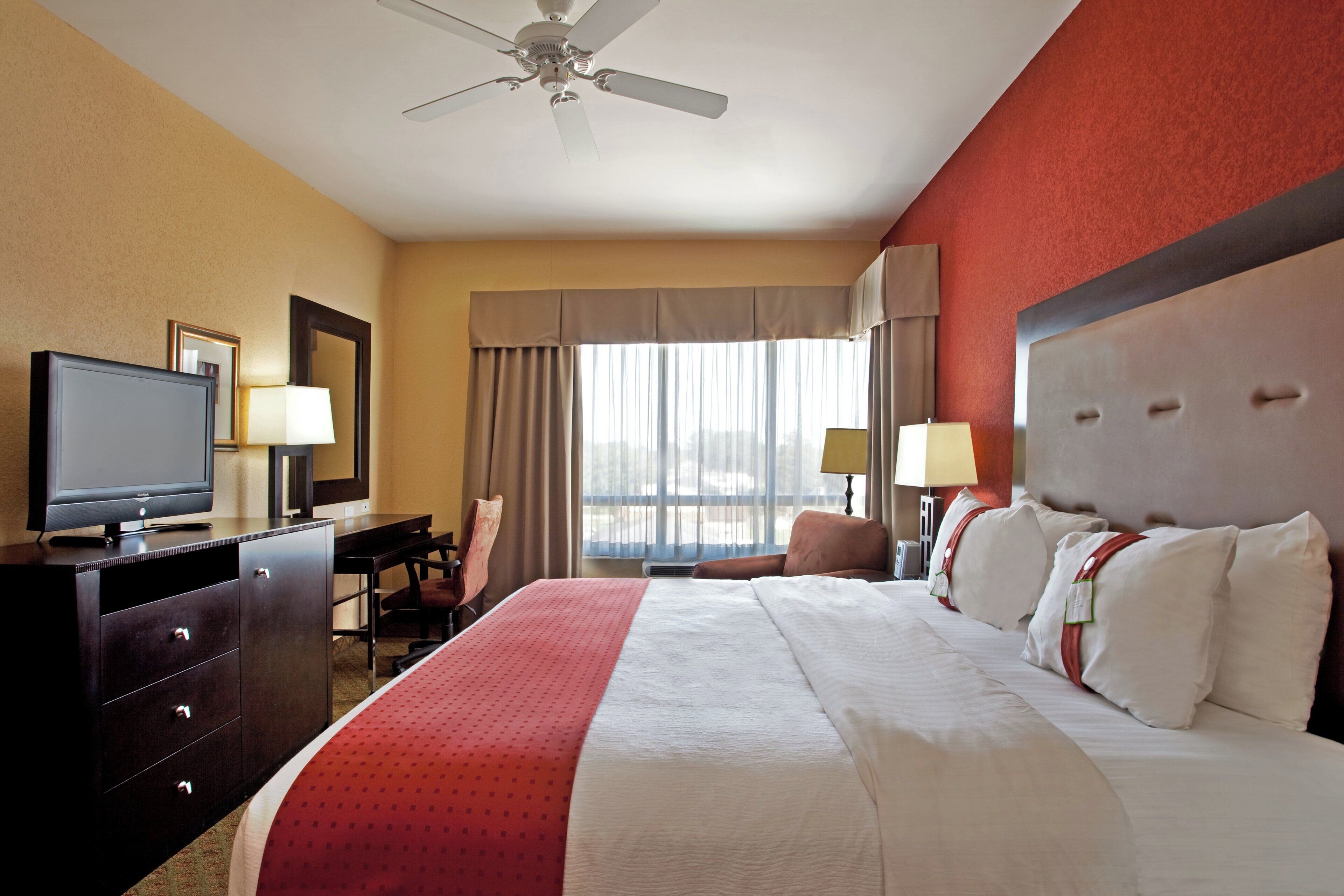 Quarto de hotel com cama, escrivaninha, televisão e ventoinha no teto.