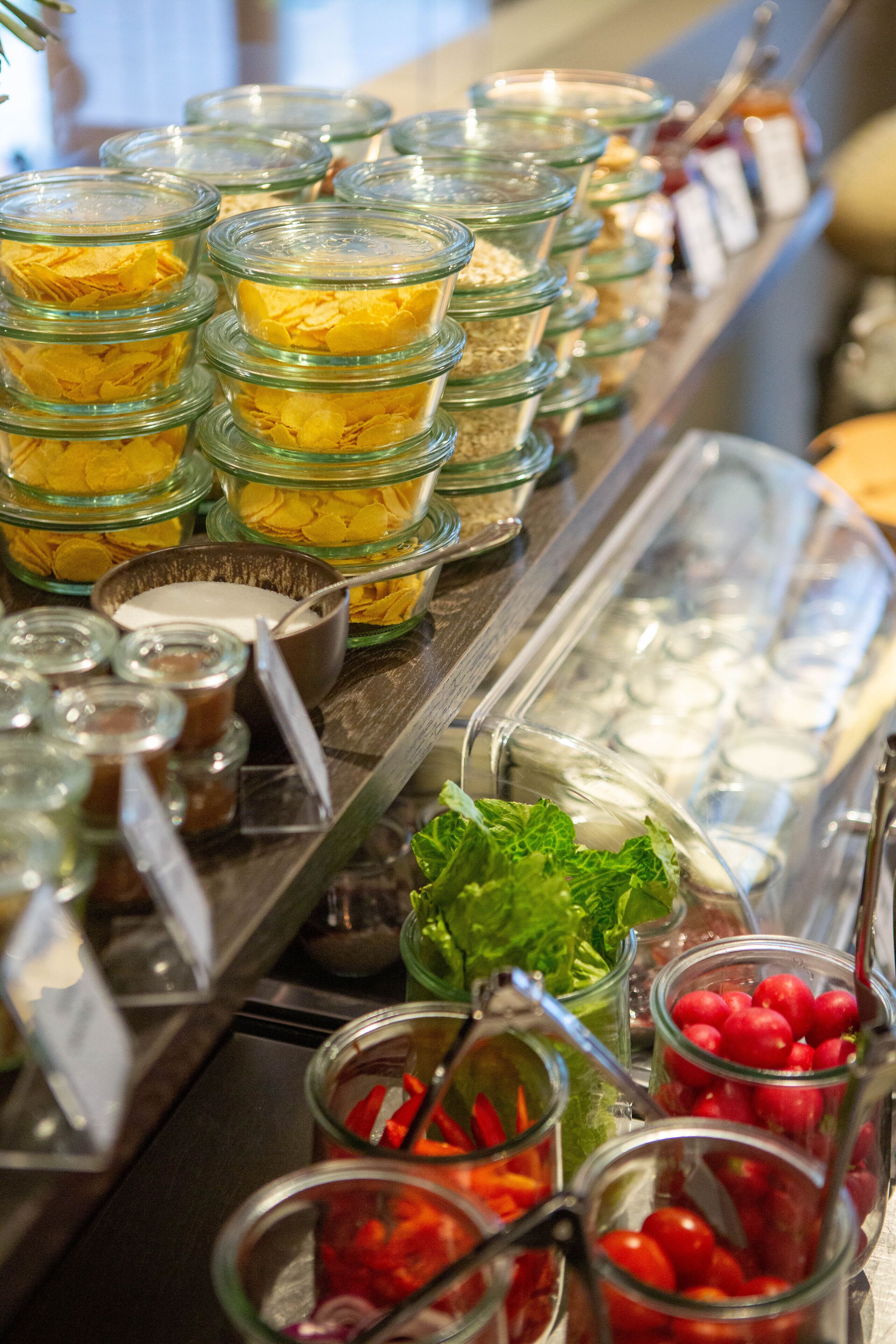 Buffet com diversos alimentos em recipientes de vidro, incluindo cereais, legumes e molhos.
