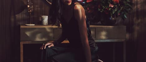 Uma mulher num vestido escuro sentada à mesa de madeira com um vaso de flores e uma lâmpada.