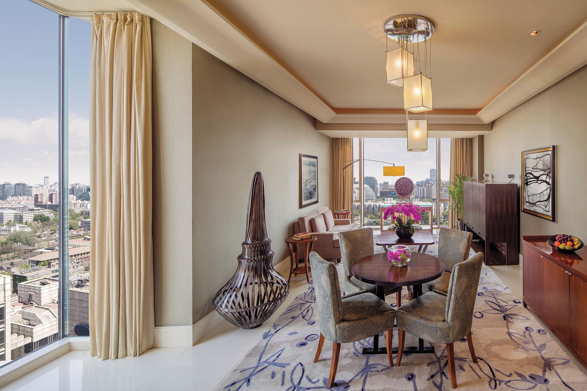 Um quarto espaçoso com vista para a cidade, uma área de jantar com mesa redonda e um grande vaso de flores.