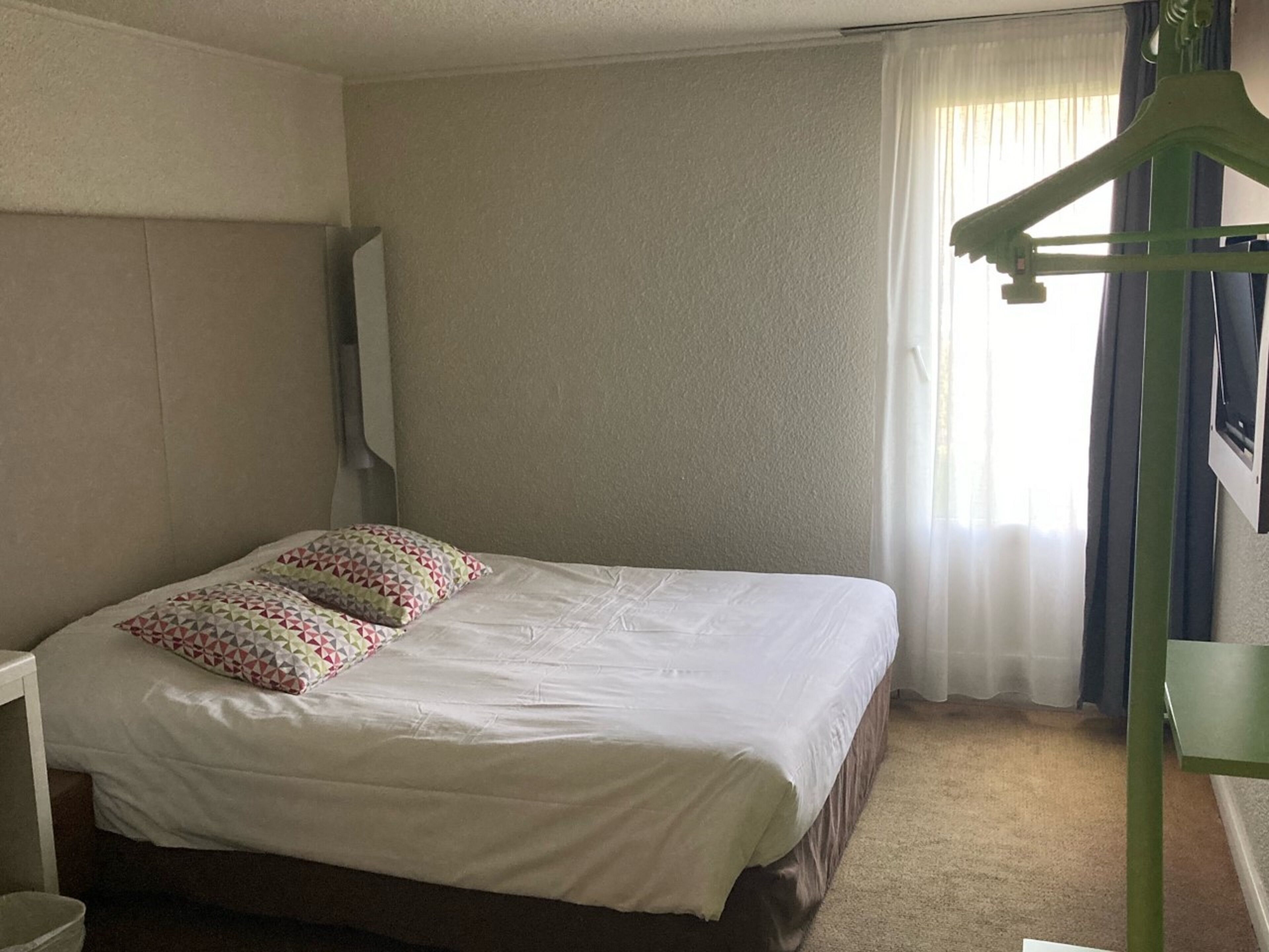 Uma cama de solteiro com almofadas estampadas, uma mesinha de cabeceira, um porta-roupas verde suspenso e uma janela com cortinas leves.