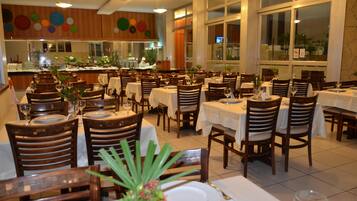 Um restaurante com várias mesas e cadeiras, uma cozinha ao fundo e grandes janelas.