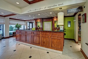 Uma área de recepção de hotel com um balcão de madeira, uma parede verde decorativa e uma placa com a inscrição 'The Pavilion Pantry'.