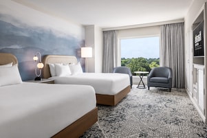Quarto de hotel com duas camas, uma escrivaninha, uma cadeira, um abajur e vista para o verde pela janela.