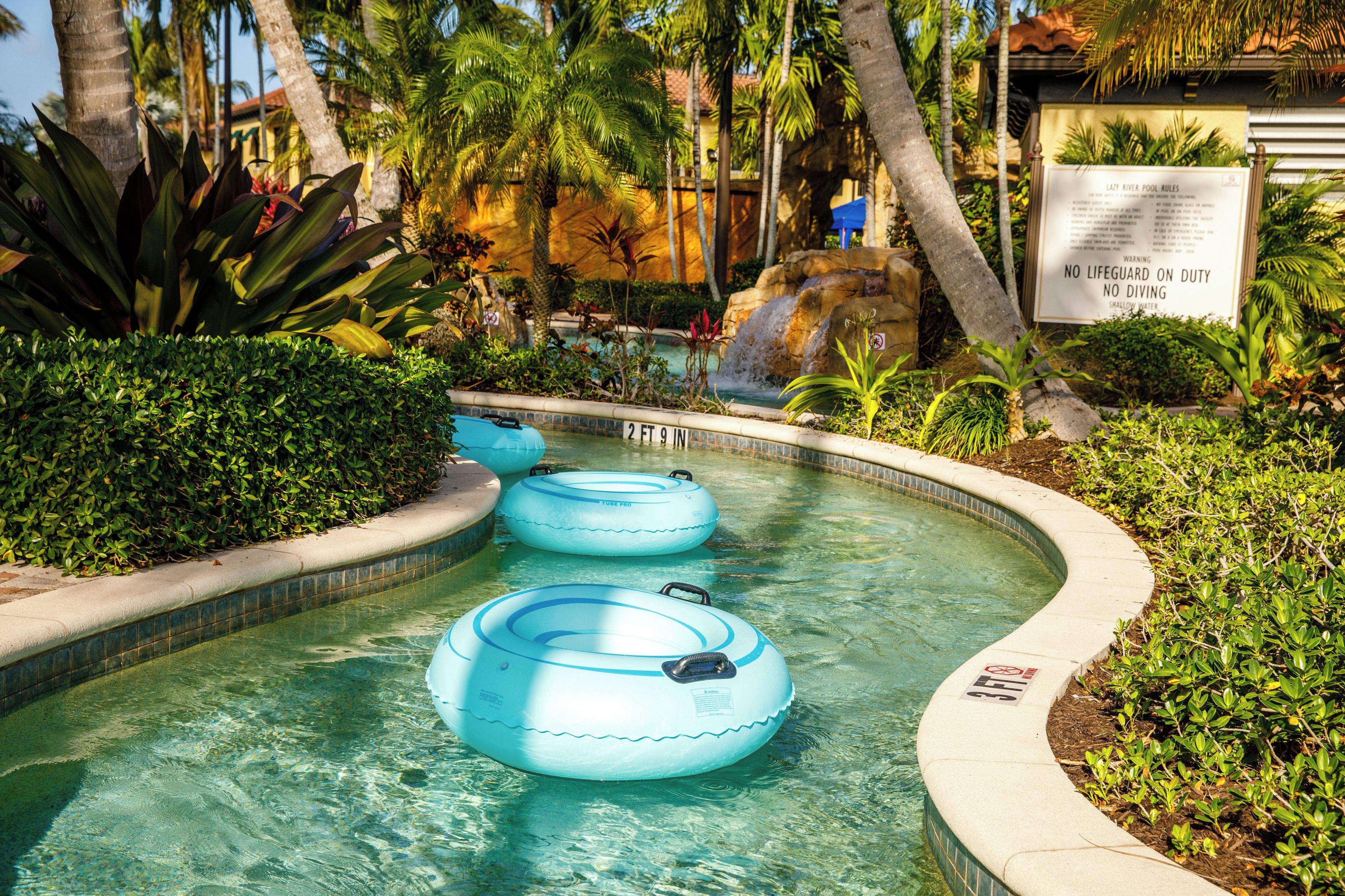 Piscina com água cristalina, rodeada por plantas tropicais e palmeiras. Há boias infláveis na piscina.