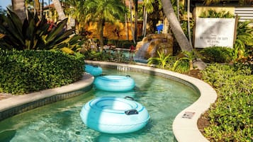 Piscina com água cristalina, rodeada por plantas tropicais e palmeiras. Há boias infláveis na piscina.