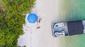 Vista aérea de uma praia com um barco atracado à direita, um guarda-sol azul e pessoas brincando na areia.