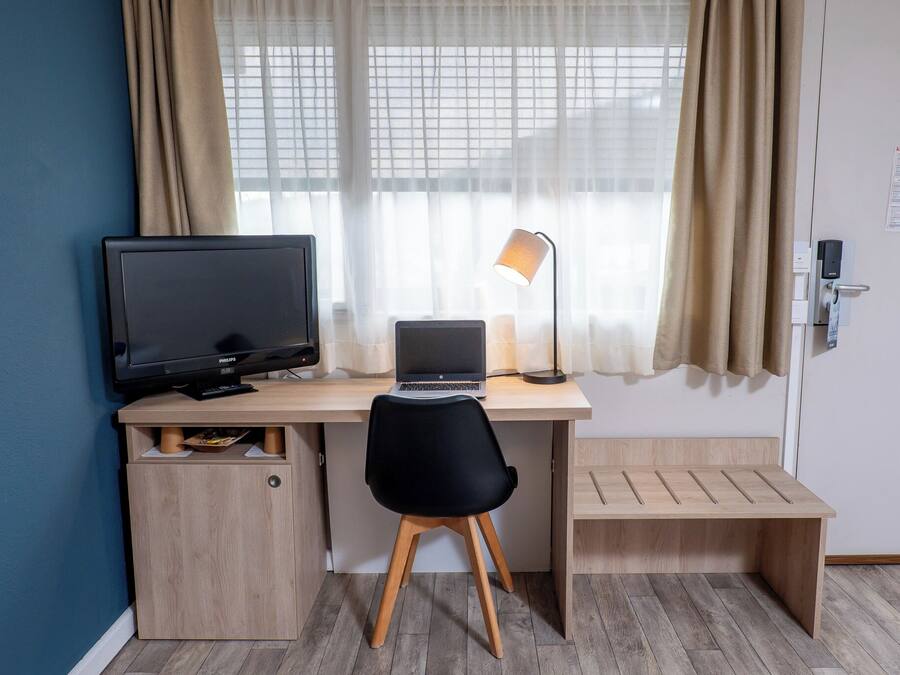 Quarto moderno com escrivaninha de madeira, cadeira preta, laptop, televisão e abajur.