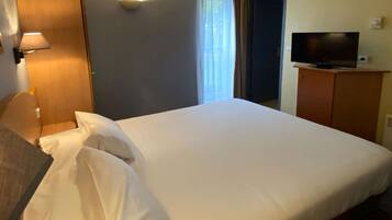 Quarto de hotel com cama bem arrumada, televisão em aparador de madeira e luminária na parede.