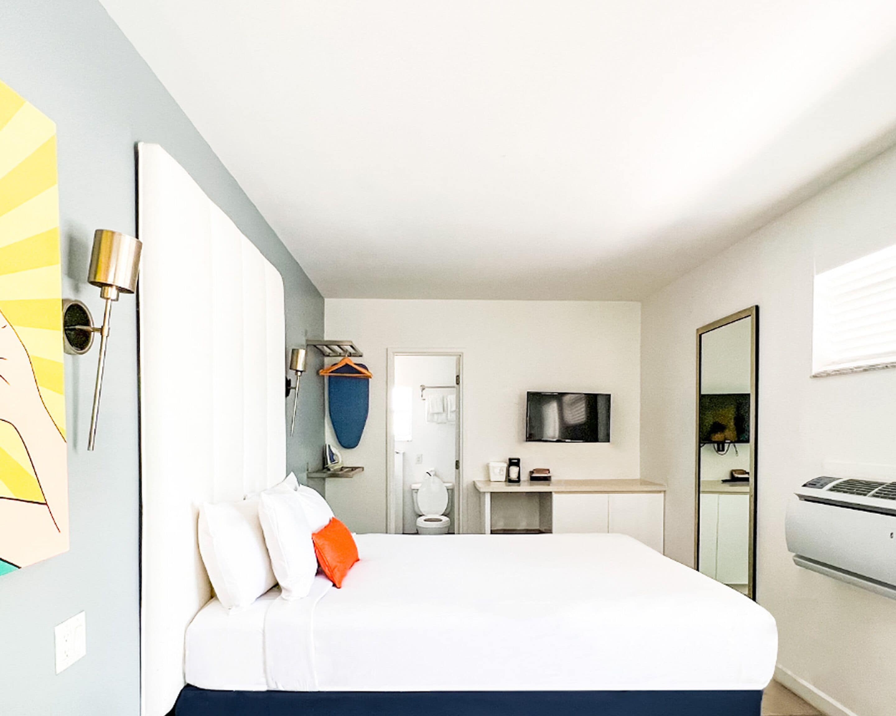 Quarto de hotel moderno com cama, escrivaninha, televisão e espelho.