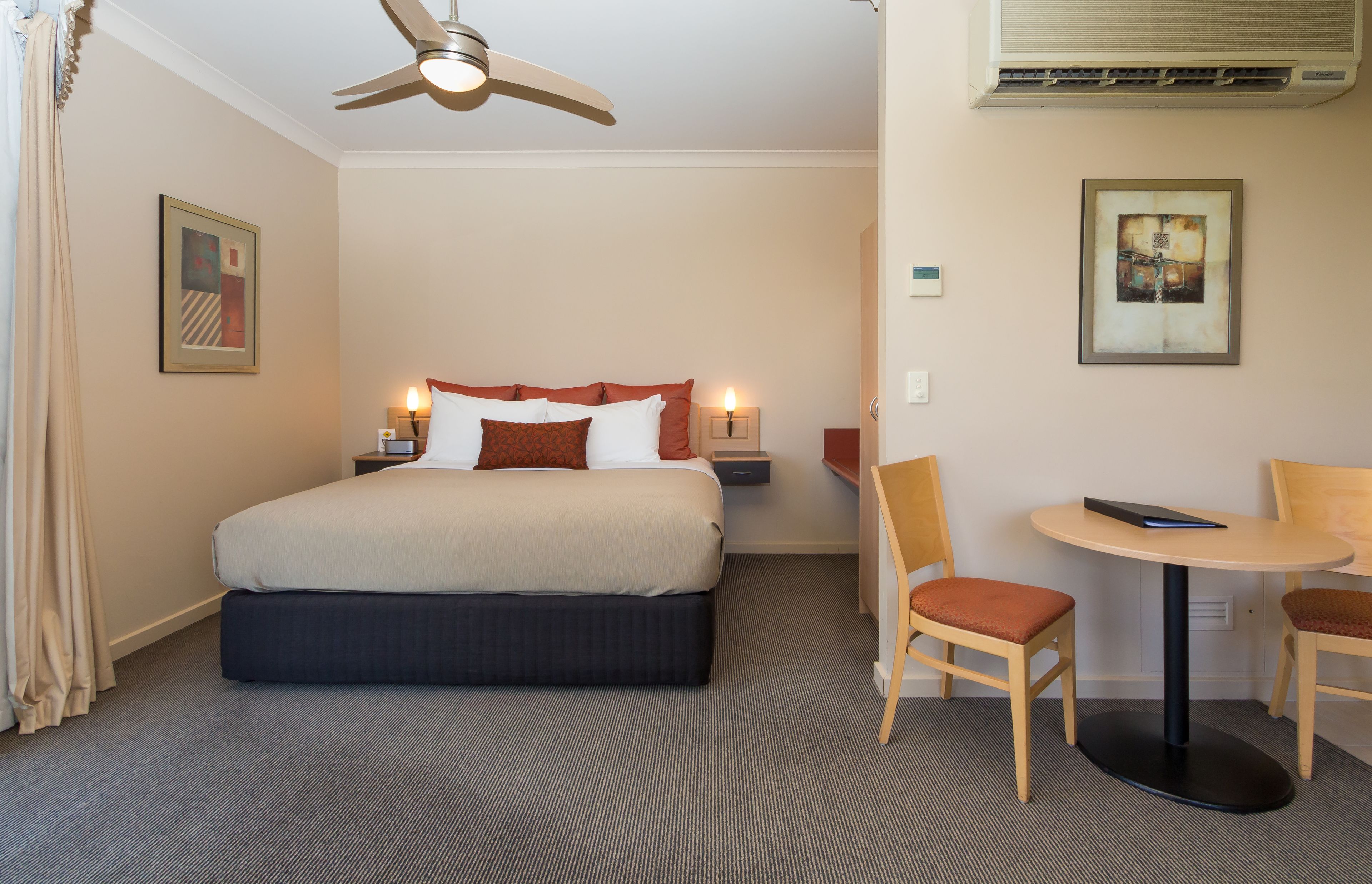 Quarto de hotel com cama, escrivaninha com cadeira, ventilador de teto e aparelho de ar condicionado.