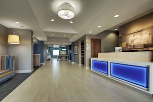 Uma área de recepção de hotel com design moderno, apresentando um balcão de atendimento com painéis azuis iluminados, um espaço de estar com sofá listrado e um corredor que leva a uma sala de jantar.