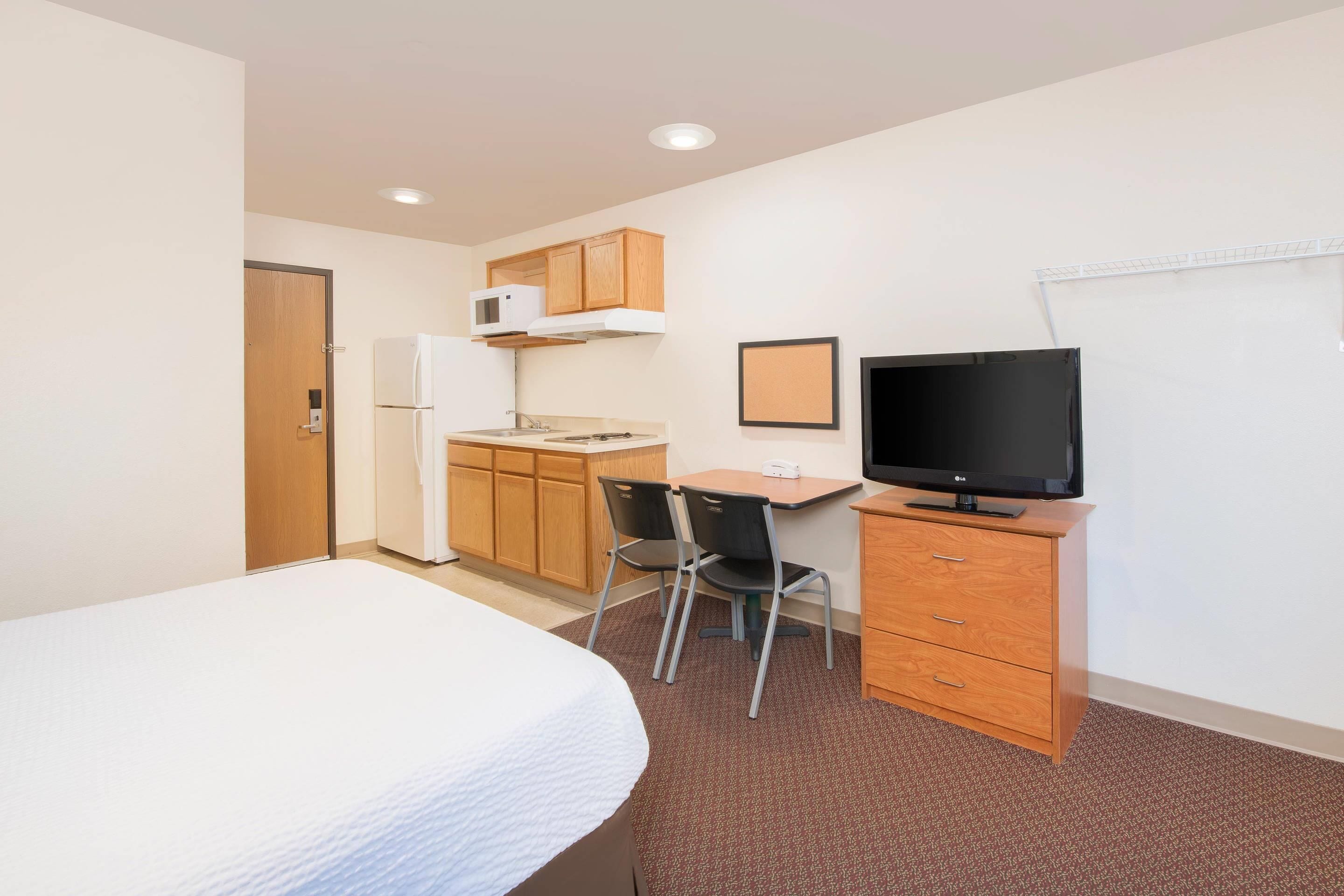 Quarto de hotel com cama, pequeno espaço de cozinha, televisão e secretária com cadeiras.