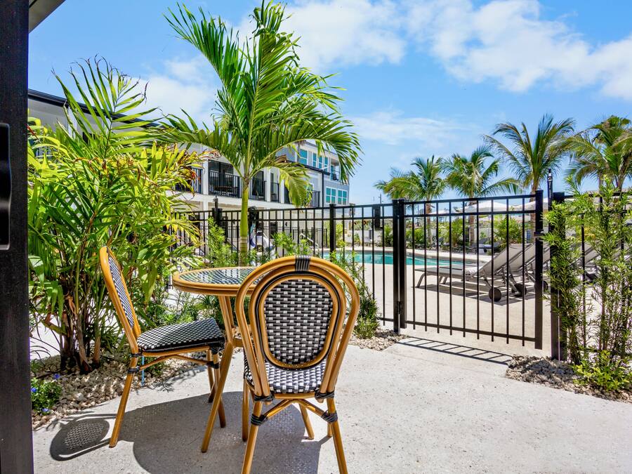 Um pátio com duas cadeiras de vime e uma mesa, com vista para uma piscina e um edifício com varandas.
