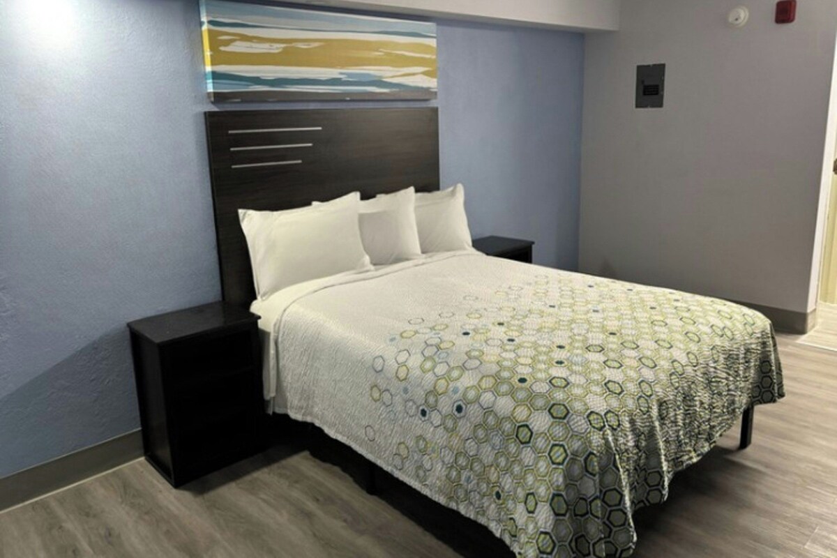 Quarto de hotel com cama, mesa de cabeceira, cabeceira com pintura e luminária fixada na parede.