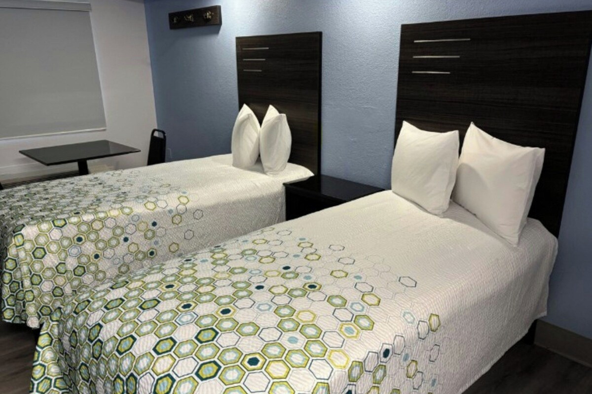 Duas camas de casal com colcha de padrão geométrico, cabeceira escura e uma pequena mesa com uma cadeira.