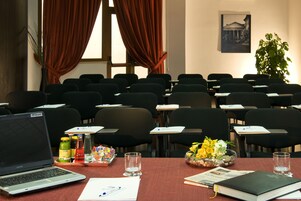 Sala de conferência com fileiras de cadeiras, um laptop, papéis e alguns petiscos sobre uma mesa.