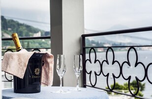 Uma garrafa de champanhe com um pano sobre uma mesa, dois copos vazios e um corrimão de varanda com vista para uma ponte e vegetação.