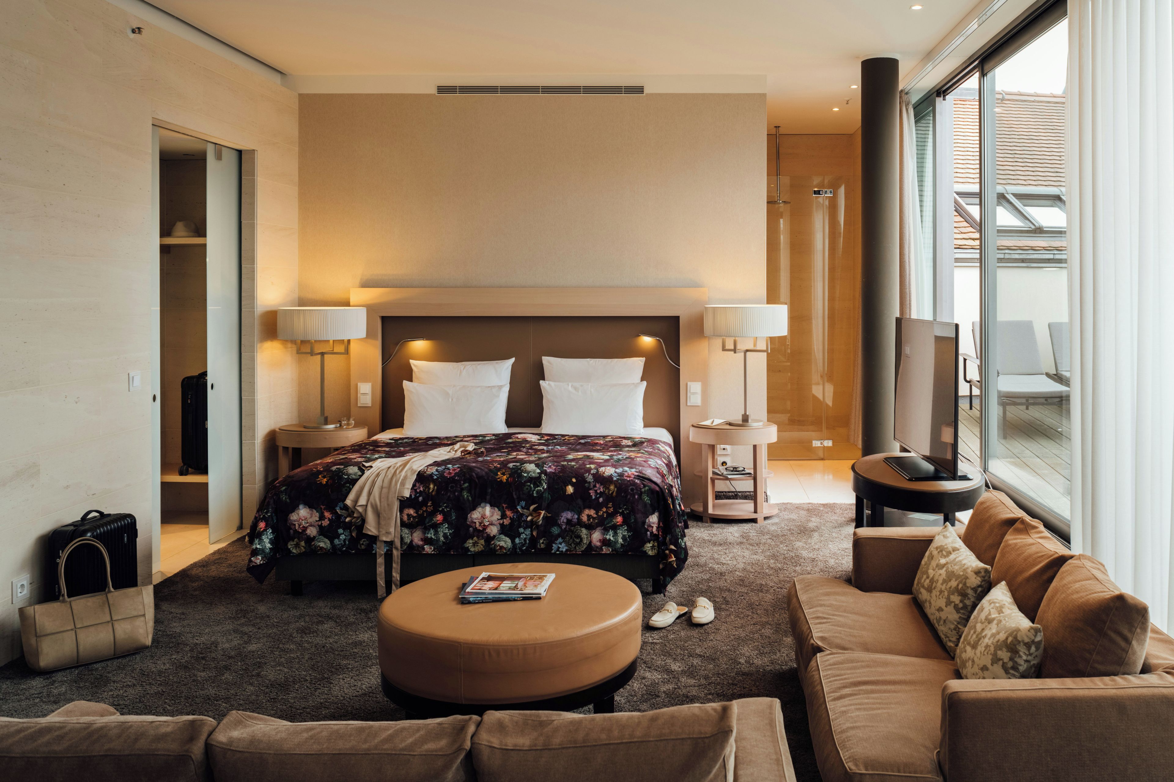 Quarto de hotel moderno com uma cama grande, duas mesas de cabeceira, um sofá e uma janela ampla.