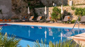 Uma piscina com espreguiçadeiras e guarda-sóis, cercada por paredes de pedra e vegetação.