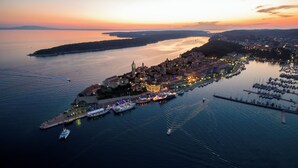 Vista aérea de uma vila costeira ao pôr do sol, com um porto, barcos e uma igreja imponente.