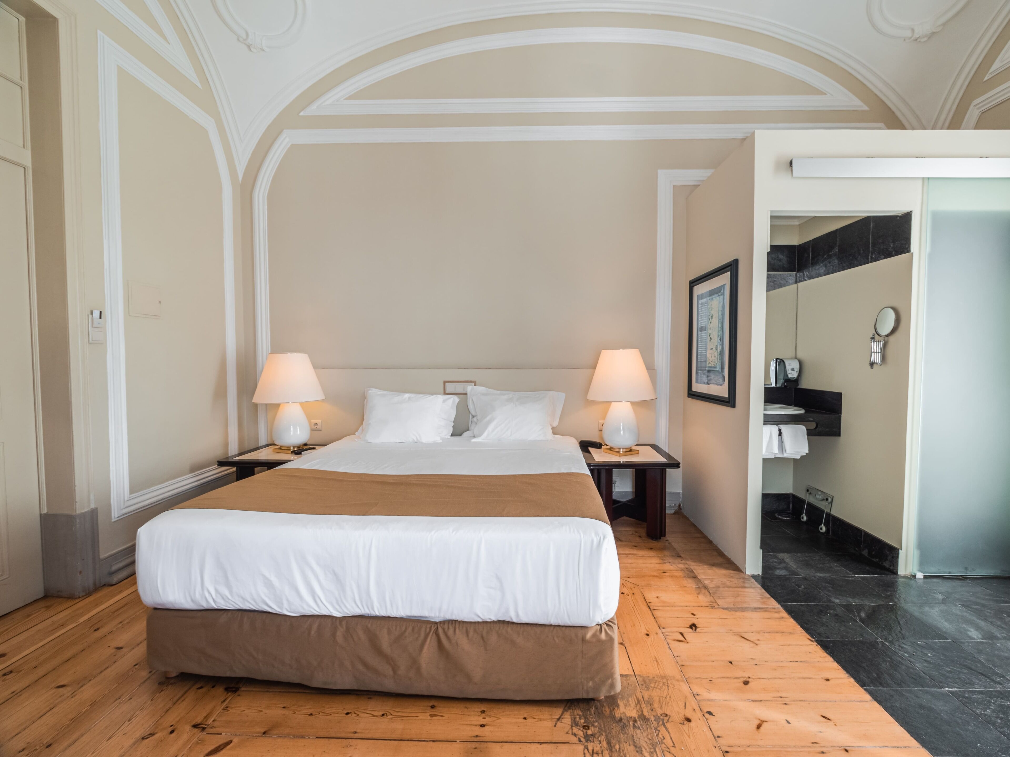 Quarto de hotel com uma cama grande, duas luminárias de cabeceira, um quadro na parede e um banheiro visível através de uma porta aberta.