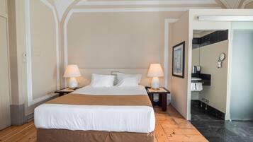 Quarto de hotel com uma cama grande, duas luminárias de cabeceira, um quadro na parede e um banheiro visível através de uma porta aberta.