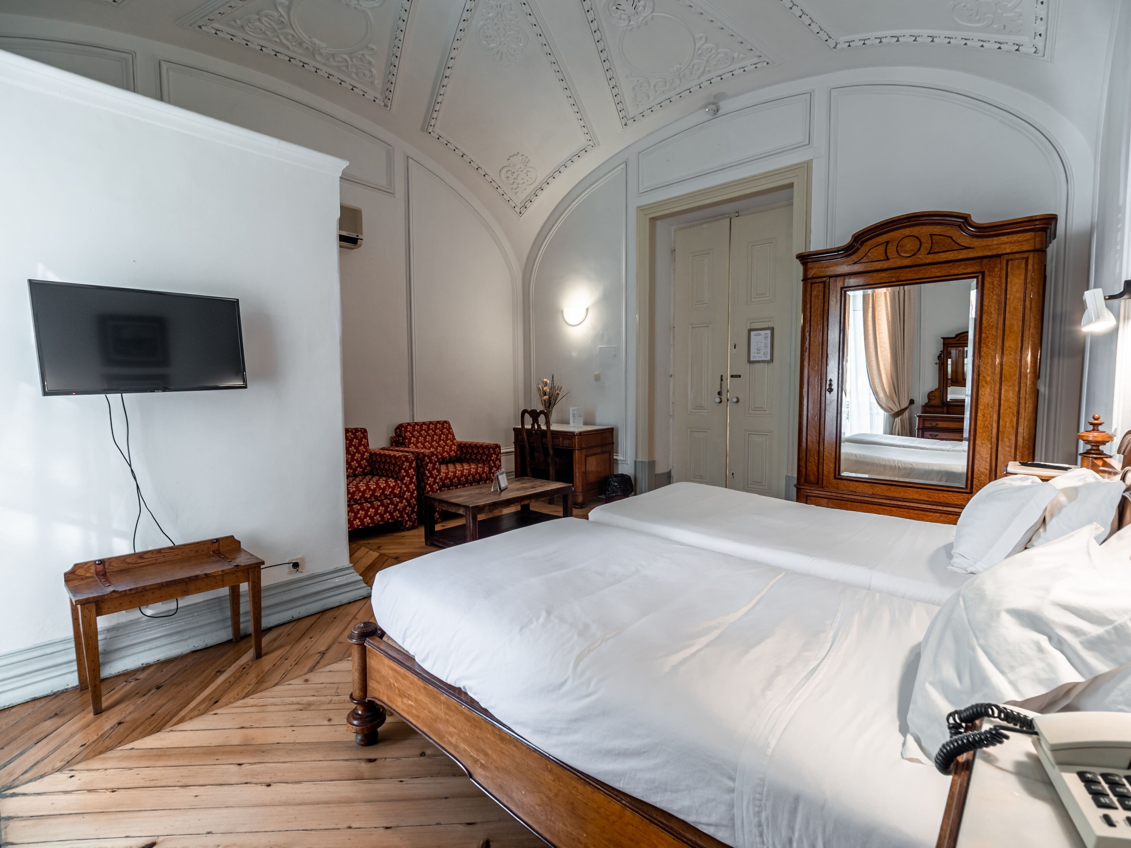 Quarto com cama grande, mesa de cabeceira de madeira, mesa auxiliar, cadeira, televisão fixada na parede e uma porta.