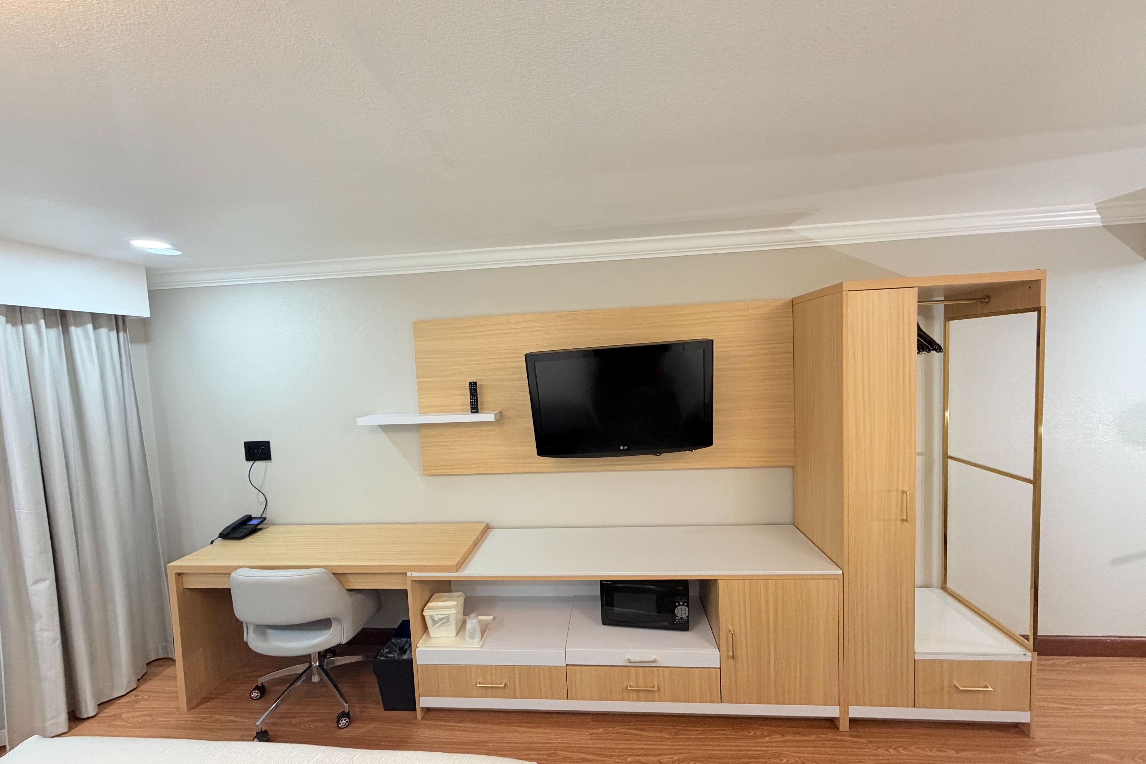 Quarto de hotel moderno com escrivaninha de madeira, TV de tela plana fixada acima, cadeira branca, prateleira pequena com micro-ondas e guarda-roupa com espelho.