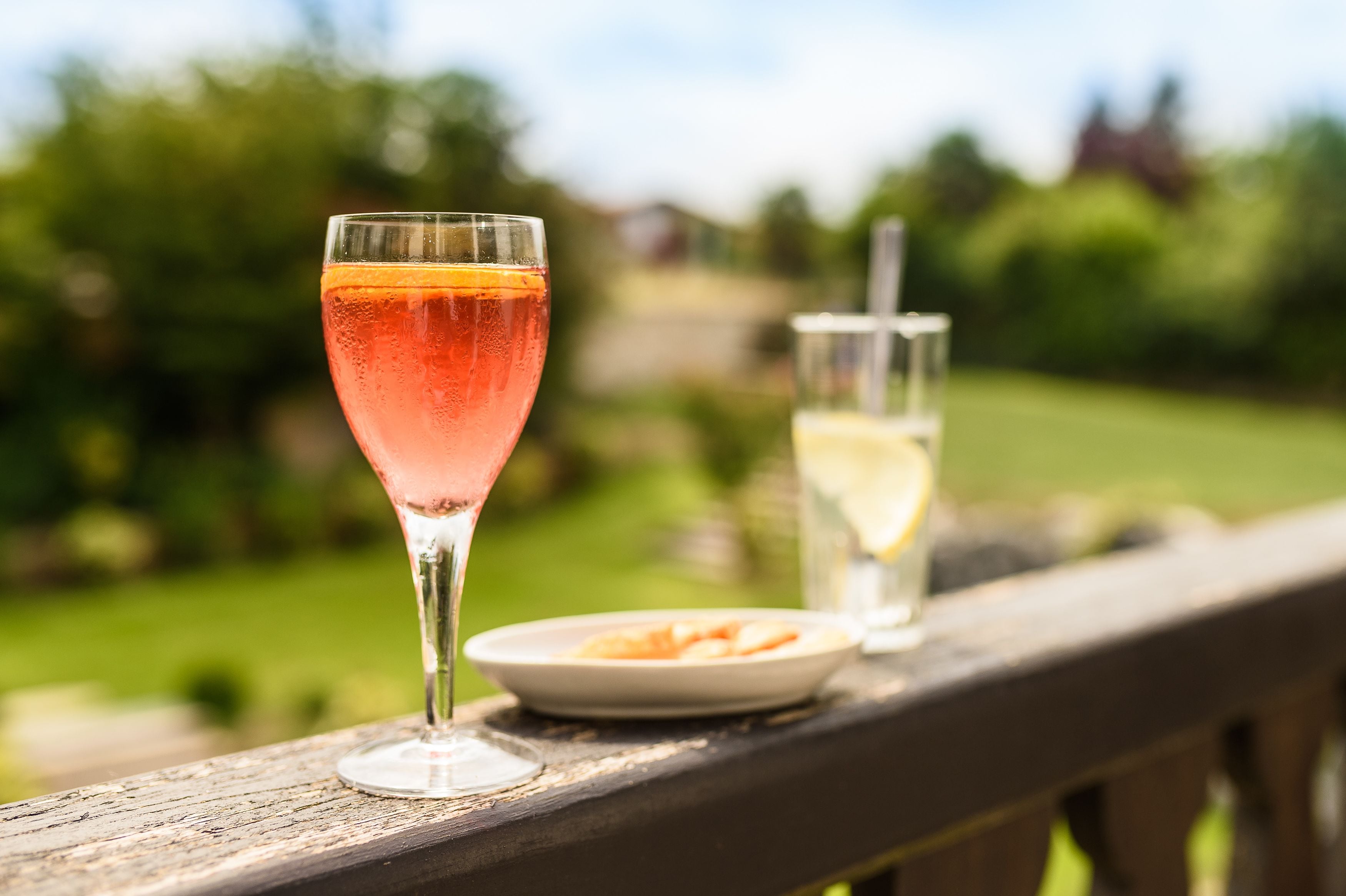 Um copo de cocktail rosa e um copo de bebida branca numa varanda de madeira, com um prato de comida e um copo de água ao fundo.