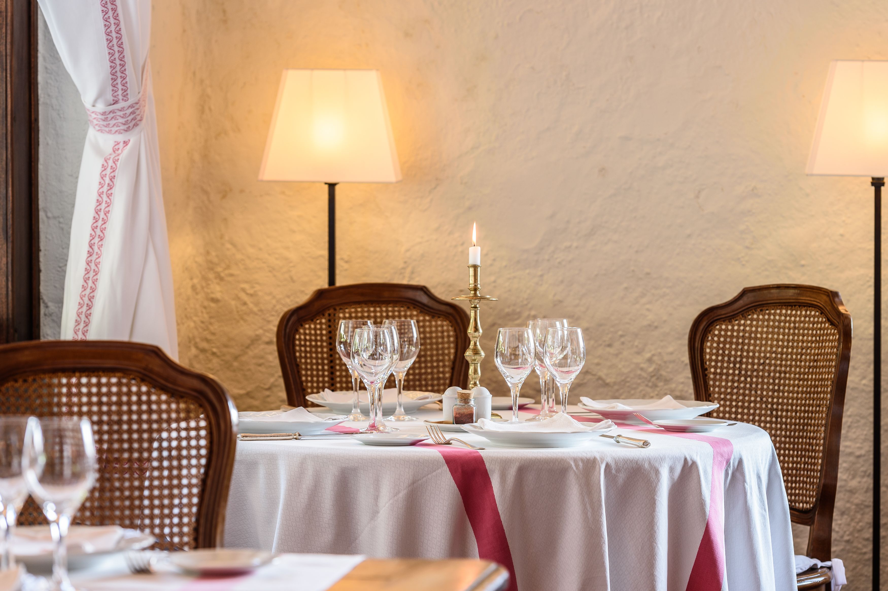 Uma mesa de jantar posta com taças de vinho, pratos e talheres, ladeada por cadeiras de vime e um abajur.