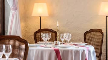 Uma mesa de jantar posta com taças de vinho, pratos e talheres, ladeada por cadeiras de vime e um abajur.