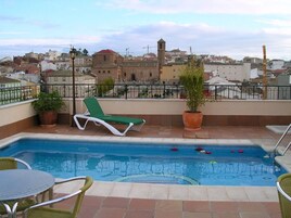 Piscina no terraço com uma espreguiçadeira verde, plantas em vasos e vista para a paisagem urbana.