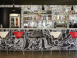 Uma área de bar com design moderno, apresentando um mural abstrato em preto e branco, diversas prateleiras com garrafas de bebidas, um refrigerador de Red Bull e banquetas de bar vermelhas e pretas.