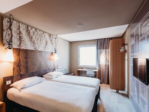 Quarto de hotel com duas camas, uma escrivaninha, uma cadeira, um armário e uma janela grande.
