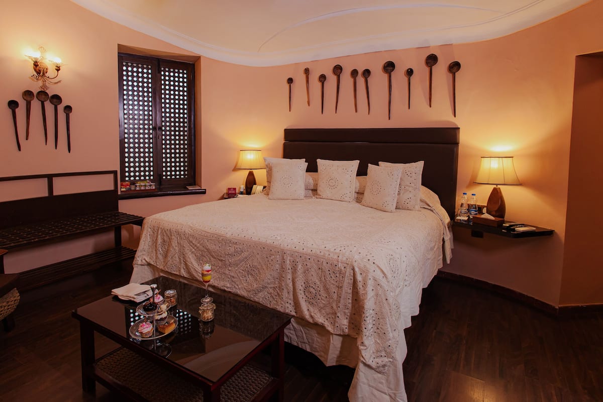 Quarto de hotel com cama grande, mesas de cabeceira com luminárias, um banco e janela com divisória decorativa.