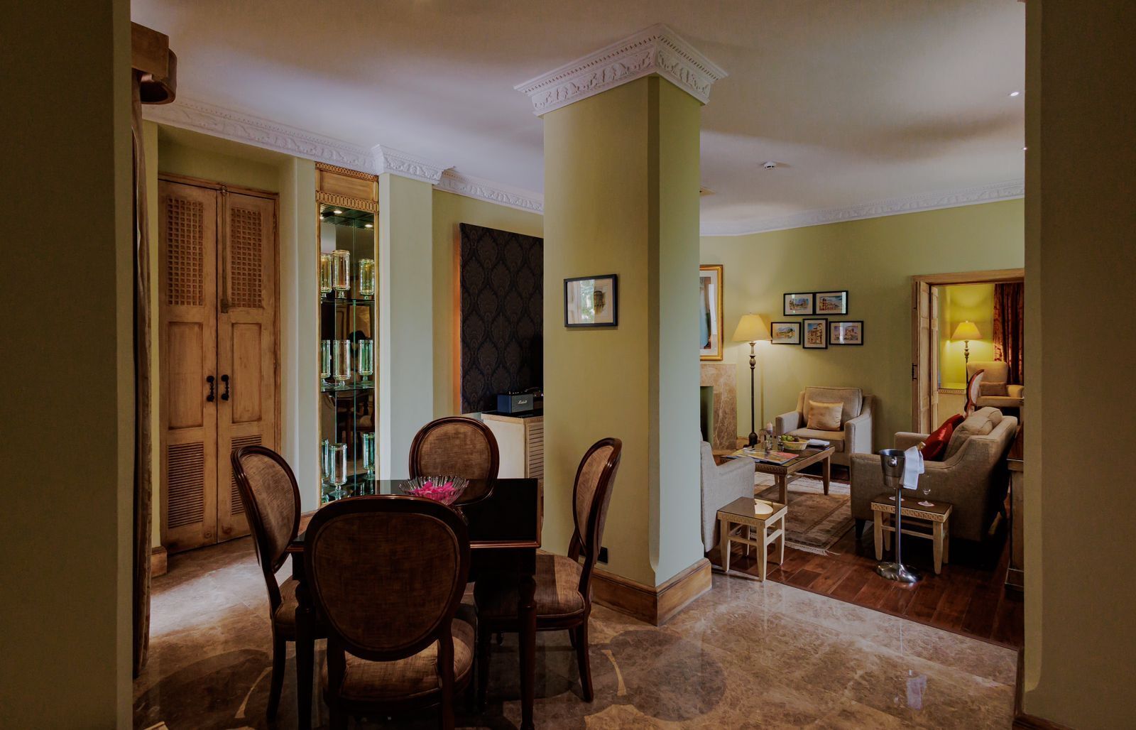 Uma sala de jantar com mesa e cadeiras, uma sala de estar com sofá e poltronas, e um corredor com portas de madeira e um armário de vidro.