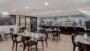 Um café limpo e moderno, com mesas e cadeiras de metal, uma lousa com saudações e um balcão de café.