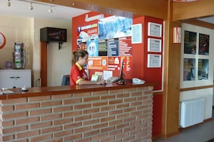 Balcão de recepção com parede vermelha, cartazes informativos, um telefone e um computador.